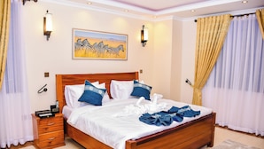 Uma cama bem arrumada com lençóis brancos e almofadas azuis, uma mesa de cabeceira de madeira com um telefone e um controle remoto, e um quadro com zebras na parede.