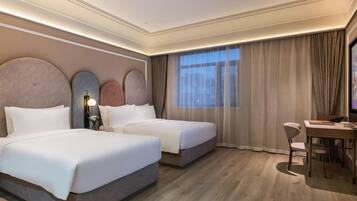 Quarto de hotel com duas camas, uma escrivaninha, uma cadeira, televisão e uma janela grande com cortinas.
