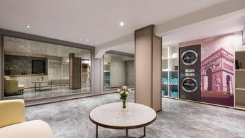 Um saguão de hotel moderno com piso de mármore, uma mesa redonda com um vaso, uma área de estar, um espelho grande e uma parede com um mural decorativo.