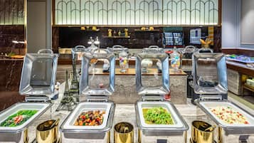 Uma área de buffet com diversos pontos de serviço, incluindo um bar de saladas, um espaço para carnes e um para massas, tudo com sinalização clara.