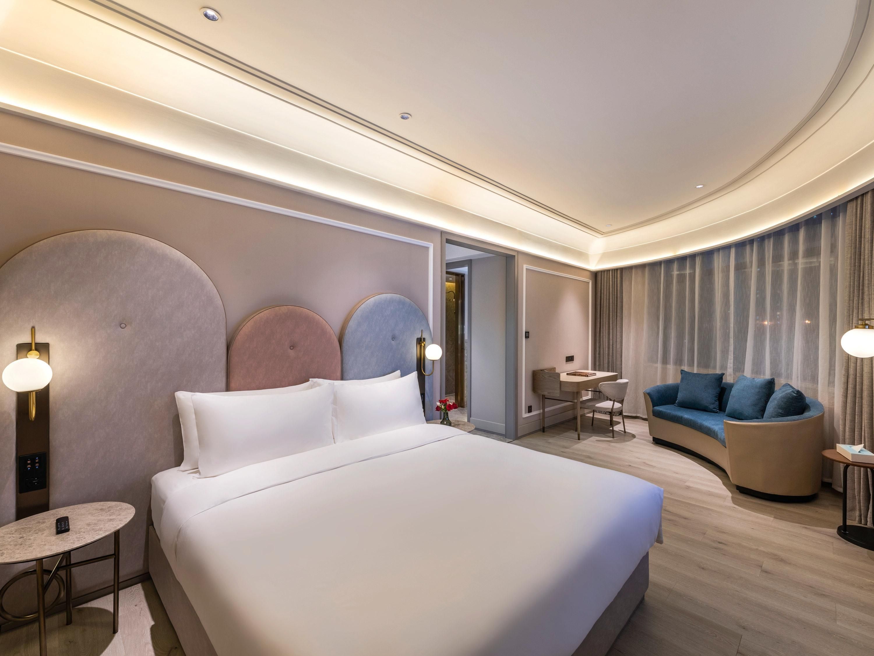Quarto de hotel moderno com uma cama grande, área de estar com sofá e cadeiras, uma mesa pequena e uma área de refeições separada.