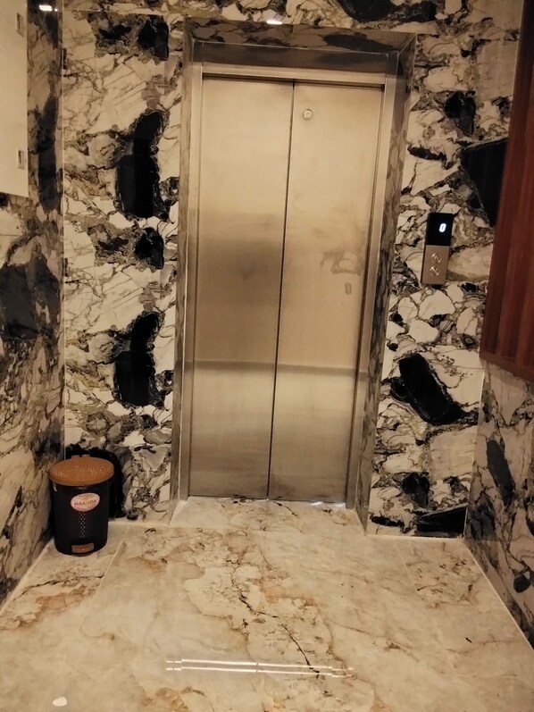 Um elevador com acabamento metálico, diante de uma parede de mármore com padrões em preto e branco.