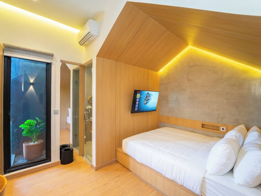 Quarto moderno com teto de madeira, uma cama grande com roupa de cama branca, uma TV montada e um vaso com planta perto da janela.