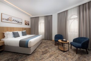 Quarto de hotel com uma cama grande, duas poltronas, uma mesinha e um quadro com vista para a cidade.