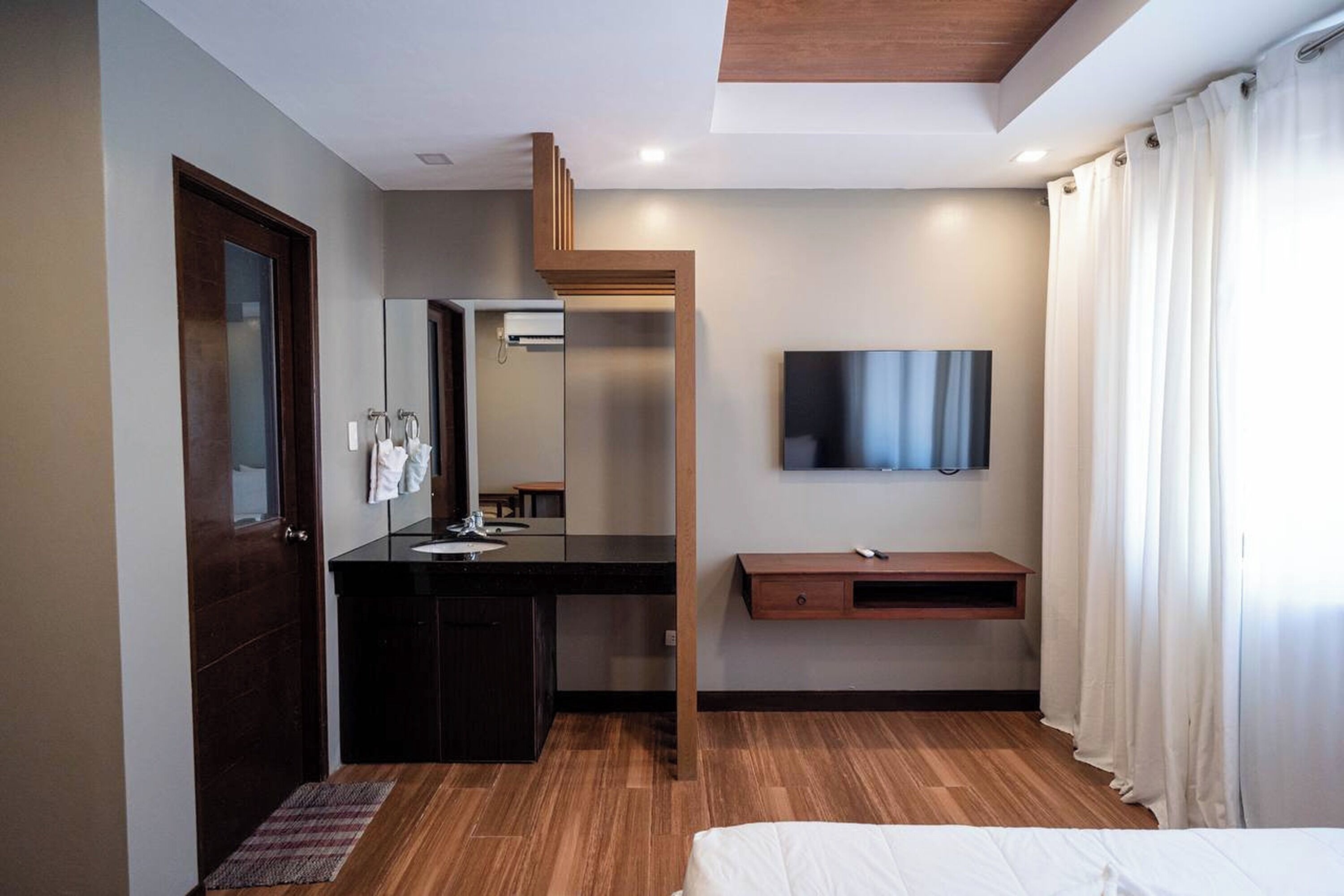 Quarto de hotel moderno com piso de madeira, escrivaninha preta com espelho, TV de tela plana na parede e cama com roupa de cama branca.