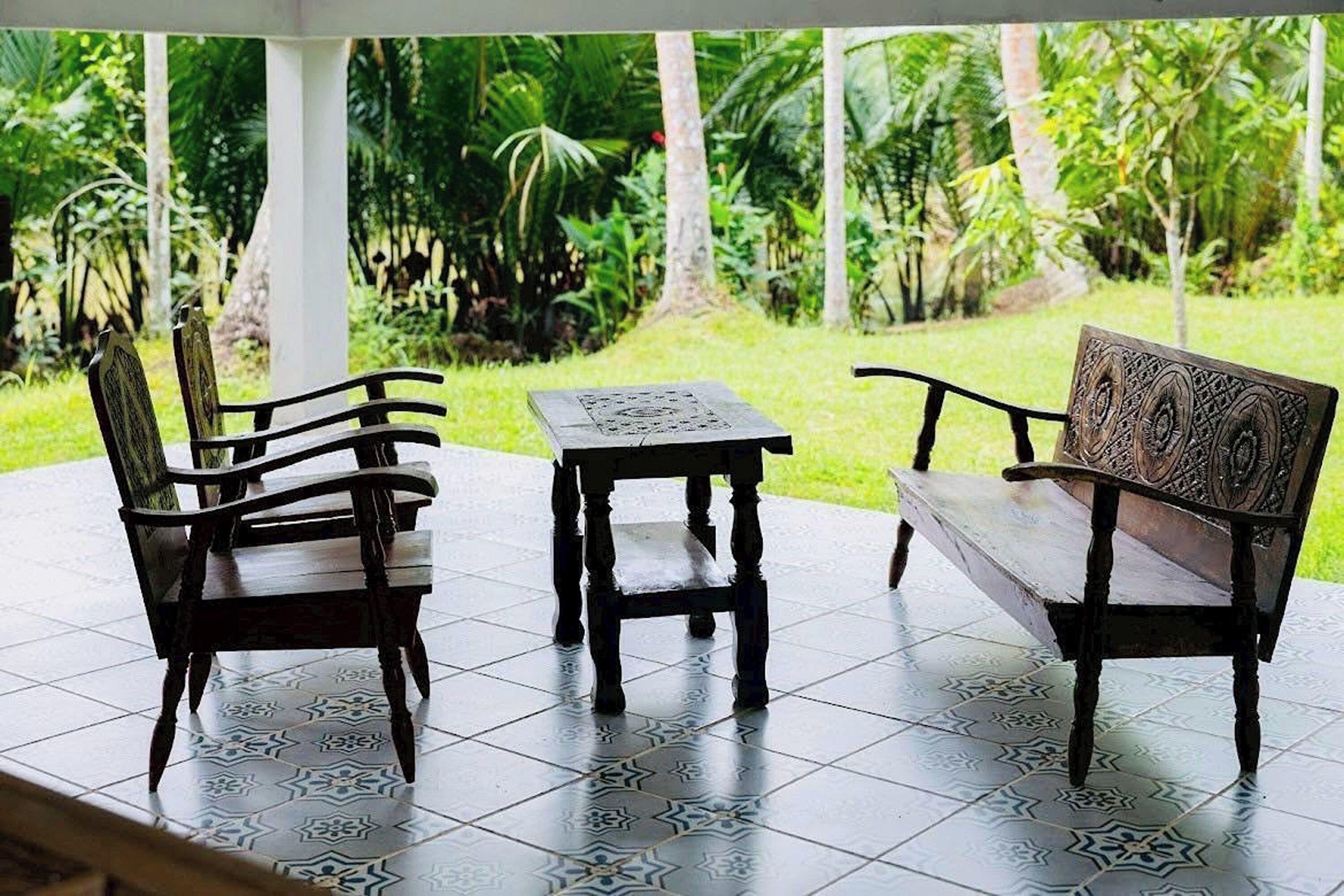 Um alpendre coberto com piso de azulejos, duas cadeiras de madeira, uma mesa pequena e um banco com detalhes entalhados.