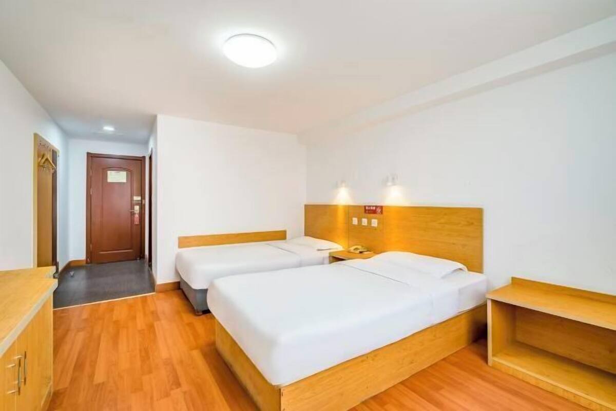 Quarto de hotel com duas camas de solteiro, cabeceira de madeira, mesa de cabeceira pequena, escrivaninha e guarda-roupa.