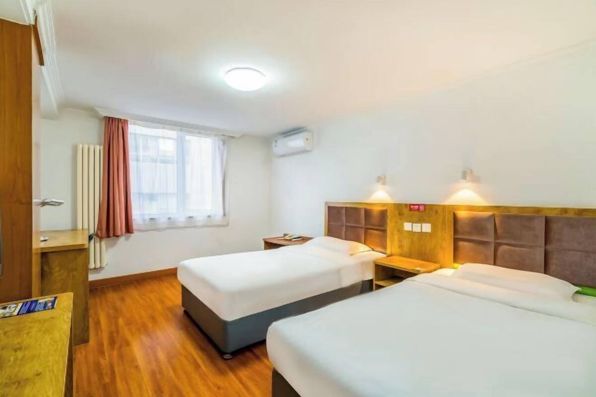 Quarto de hotel com duas camas, piso de madeira, janela com cortinas, unidade de ar condicionado e mesa de cabeceira com luminária.