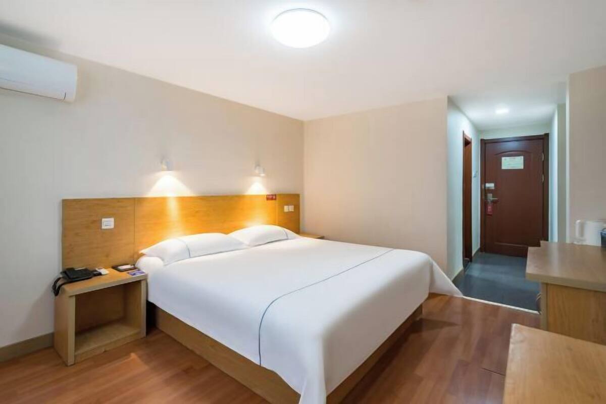 Quarto de hotel com cabeceira de madeira, duas luminárias de apoio, mesa de cabeceira de madeira, cama com roupa de cama branca, escrivaninha de madeira e porta que dá para um corredor.