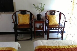 Quarto de hotel com duas cadeiras de madeira com almofadas amarelas, uma mesa pequena com uma planta em vaso e uma cama com colcha branca e vermelha.