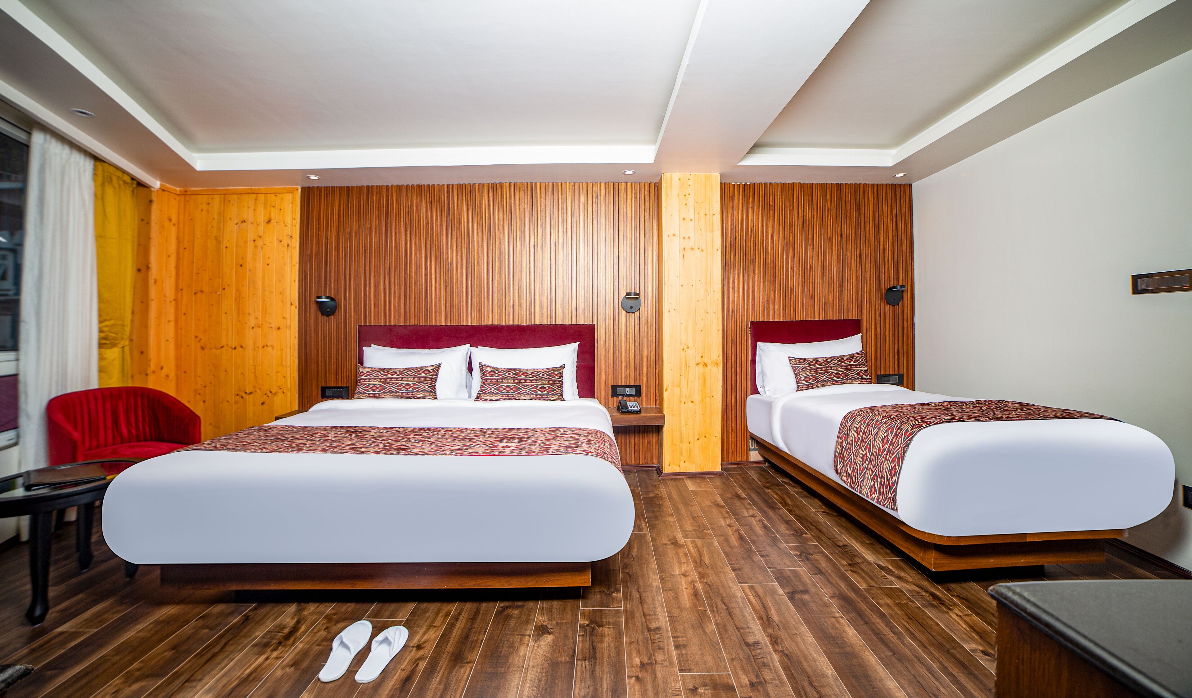 Quarto de hotel com duas camas, paredes de painel de madeira, uma cadeira vermelha, uma escrivaninha e um par de chinelos no chão.