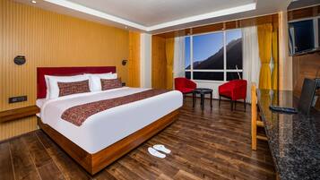 Quarto de hotel com uma cama grande, uma mesa pequena, duas cadeiras vermelhas, uma televisão e uma janela com vista para as montanhas.