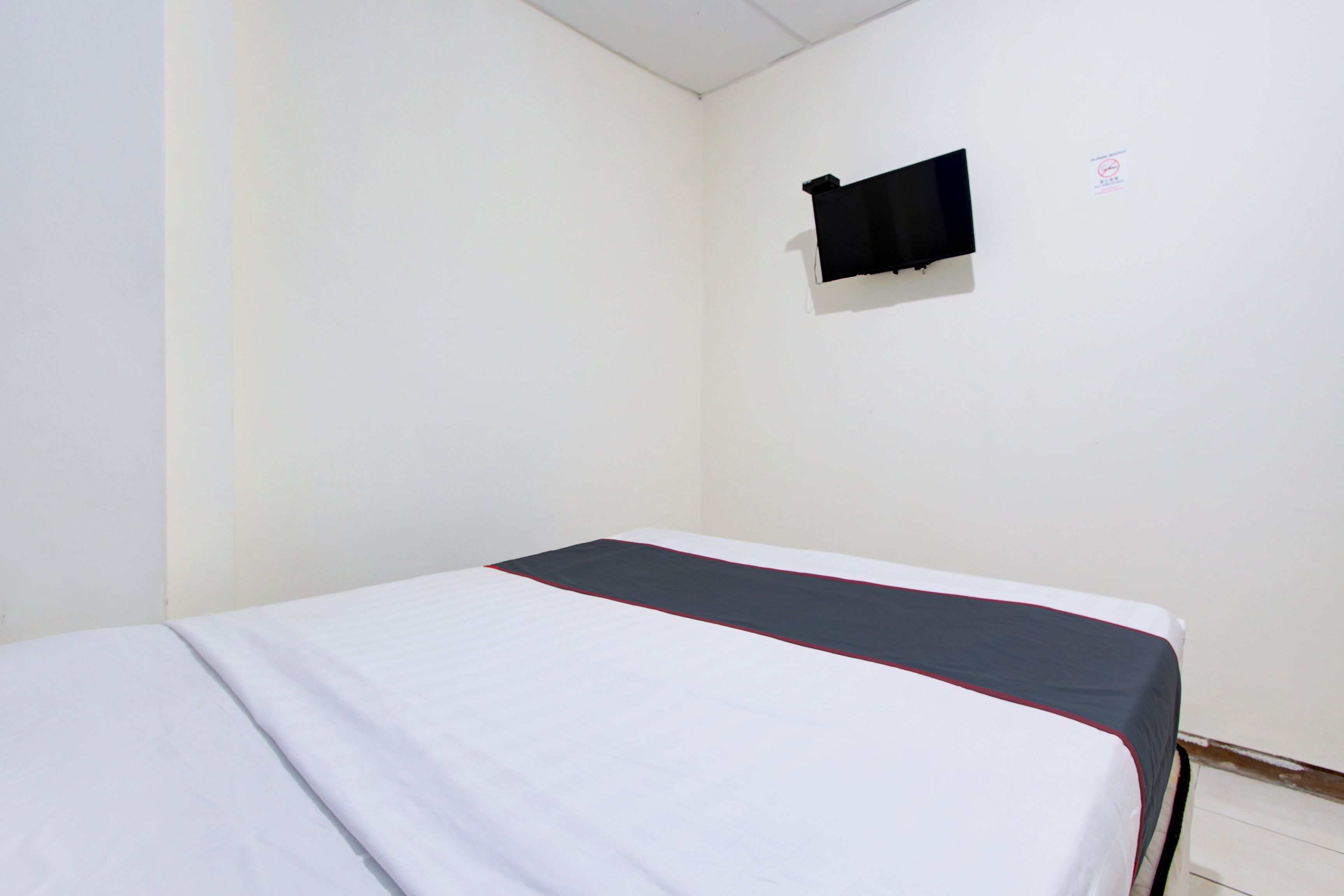 Uma cama de solteiro com colcha branca e tons escuros em um quarto com paredes brancas e televisão na parede.