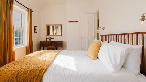 Quarto com cama, mesa de cabeceira, cadeira, escrivaninha e janela com cortinas.