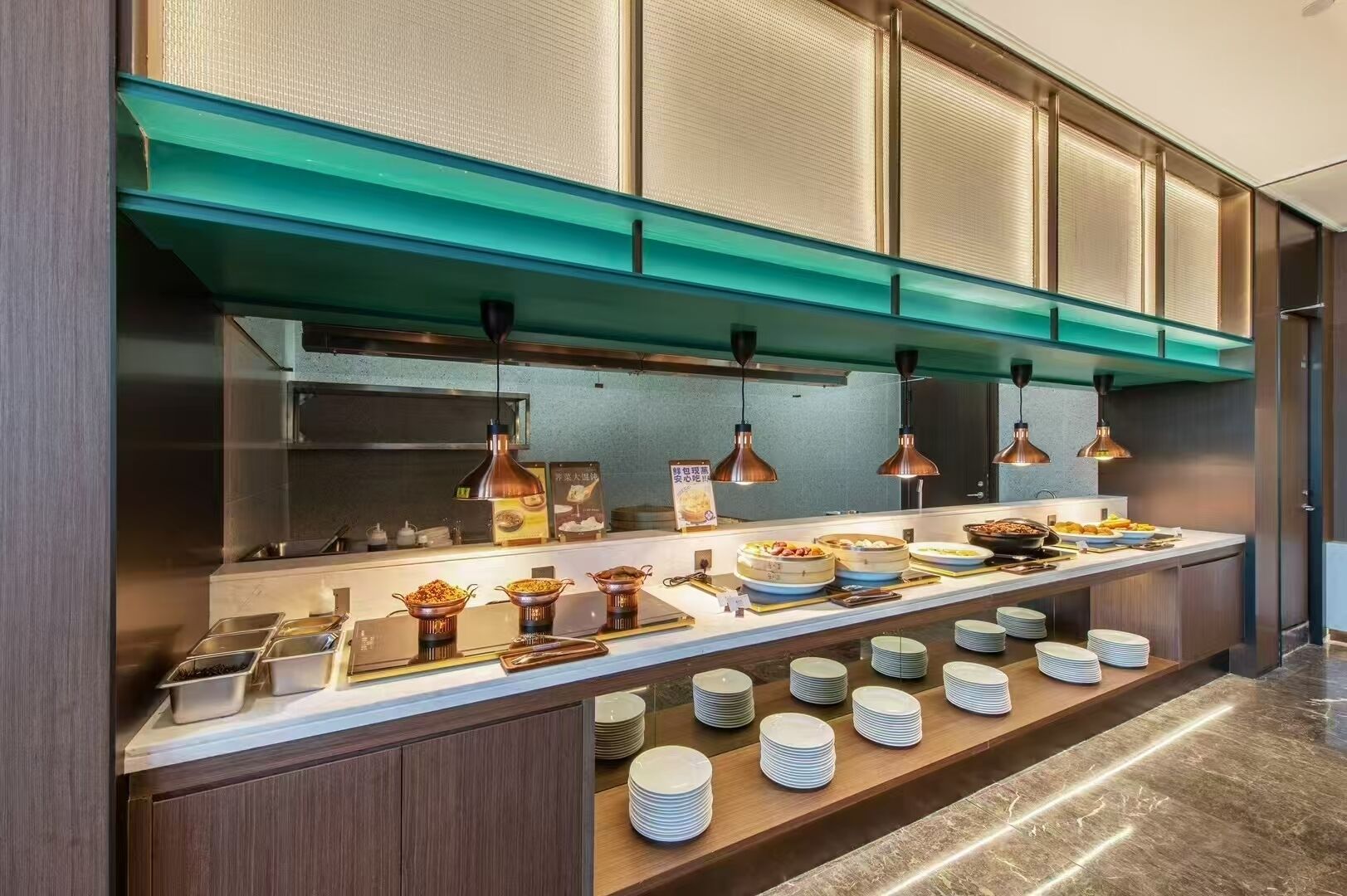 Disposição de buffet com diversos alimentos, utensílios, balcão com pratos empilhados e iluminação pendente.