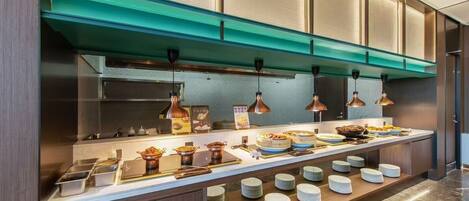 Disposição de buffet com diversos alimentos, utensílios, balcão com pratos empilhados e iluminação pendente.