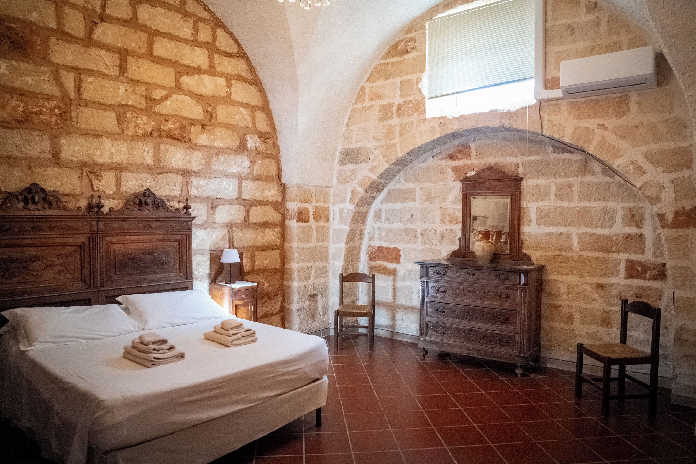 Quarto com parede de pedra, uma cama, cômoda e espelho.