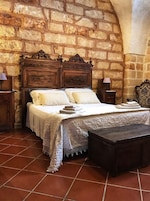 Quarto com parede de pedra, cama de madeira com cabeceira entalhada, duas mesas de cabeceira com luminárias, um pequeno banco e piso ladrilhado.