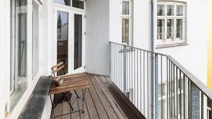 Uma varanda com mesa e cadeiras de madeira, guarda-corpo de metal e porta que dá para o interior.