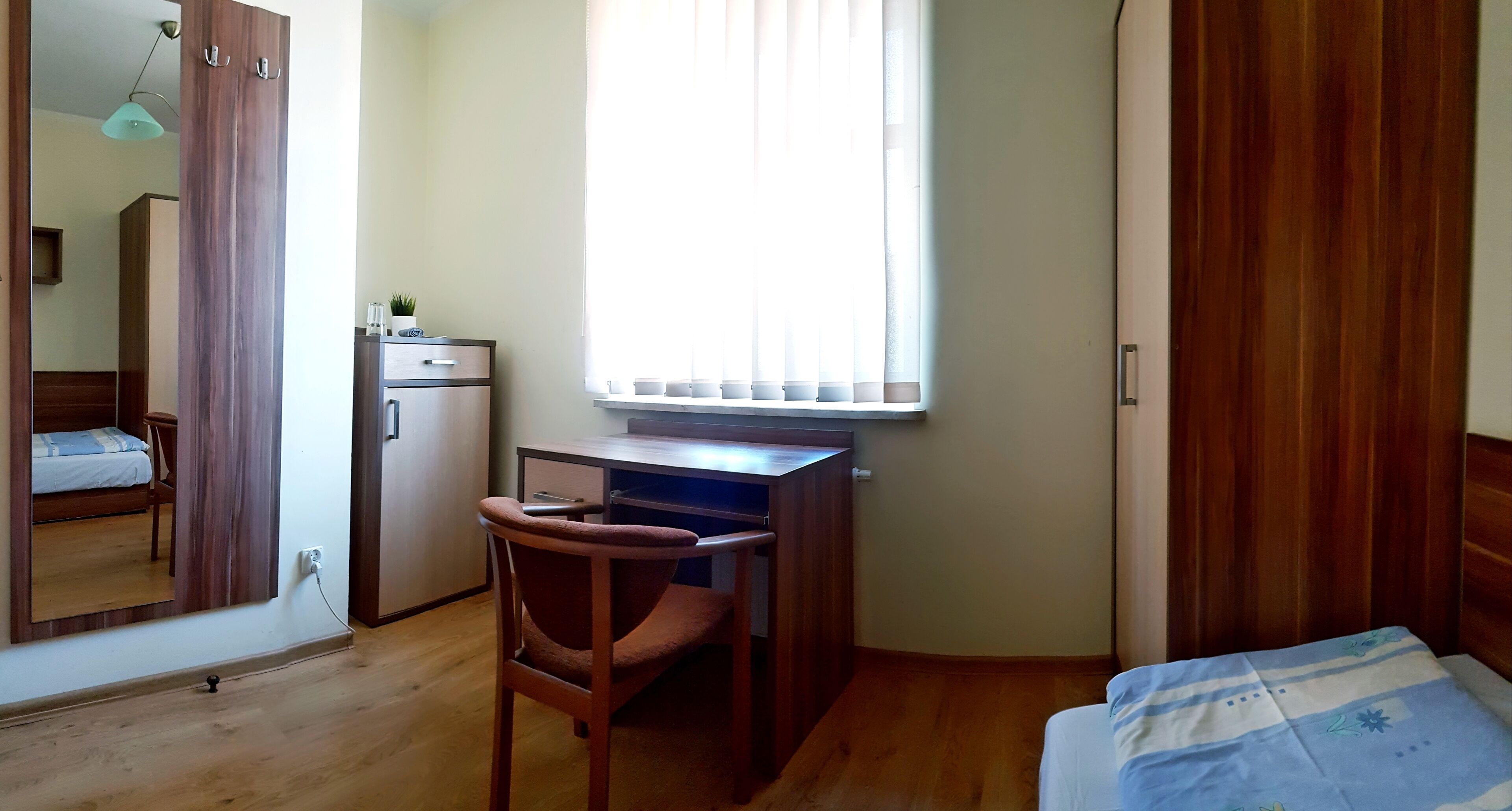 Quarto com escrivaninha de madeira, uma cadeira, cama com colcha listrada, janela com persianas e guarda-roupa.