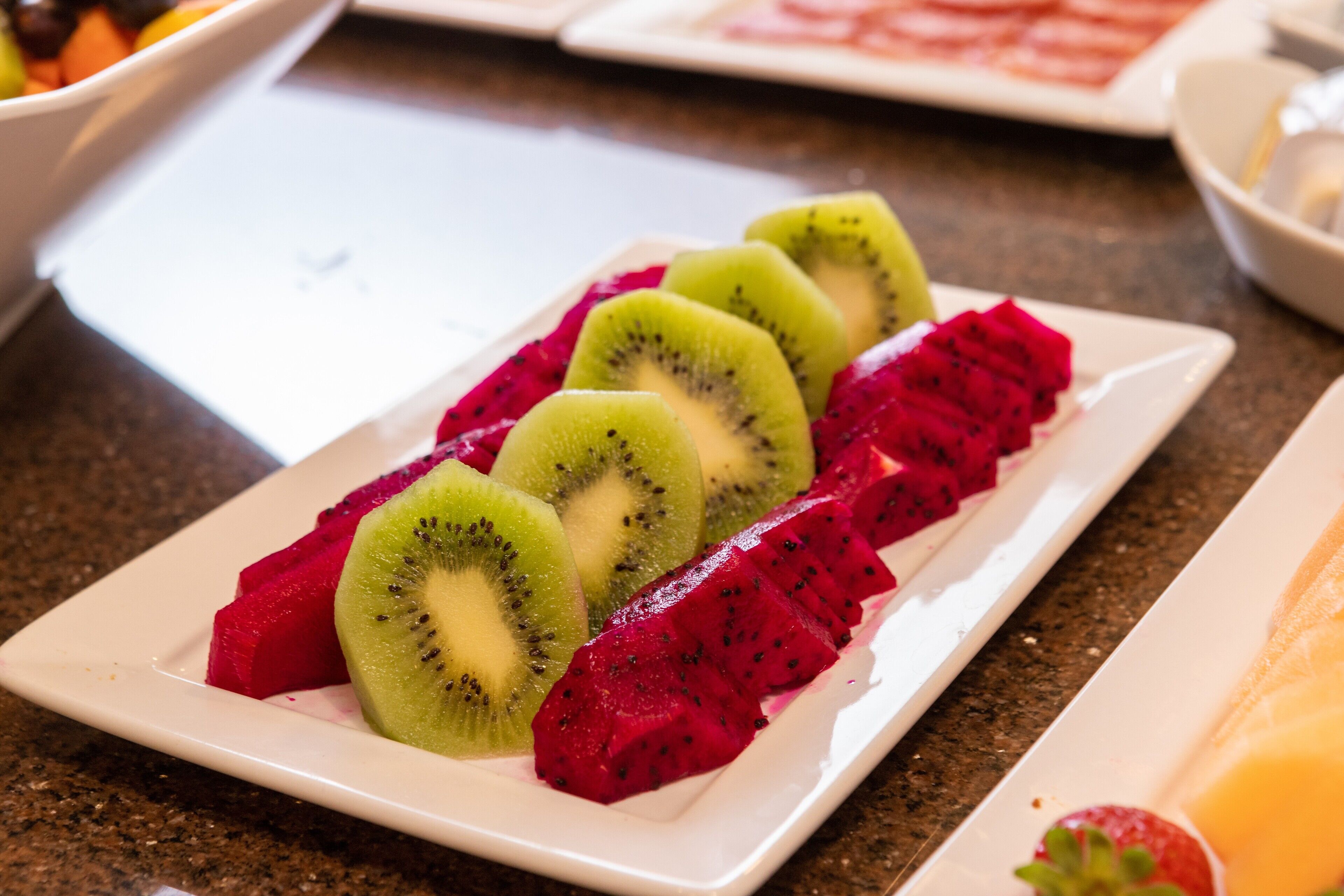 Prato branco retangular com kiwi fatiado e pitaya sobre bancada de granito.