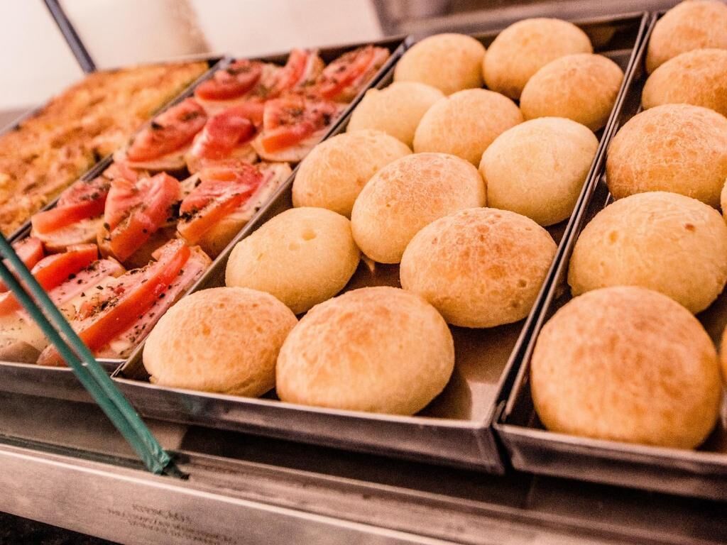 Buffet com diversos pães e produtos de panificação, incluindo mini pães e um prato com tomate e carne fatiados.