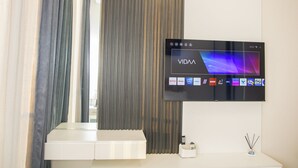Quarto de hotel moderno com uma TV de tela plana montada, exibindo a marca VIDAA, um console de mesa branco e um controle remoto sobre o console.