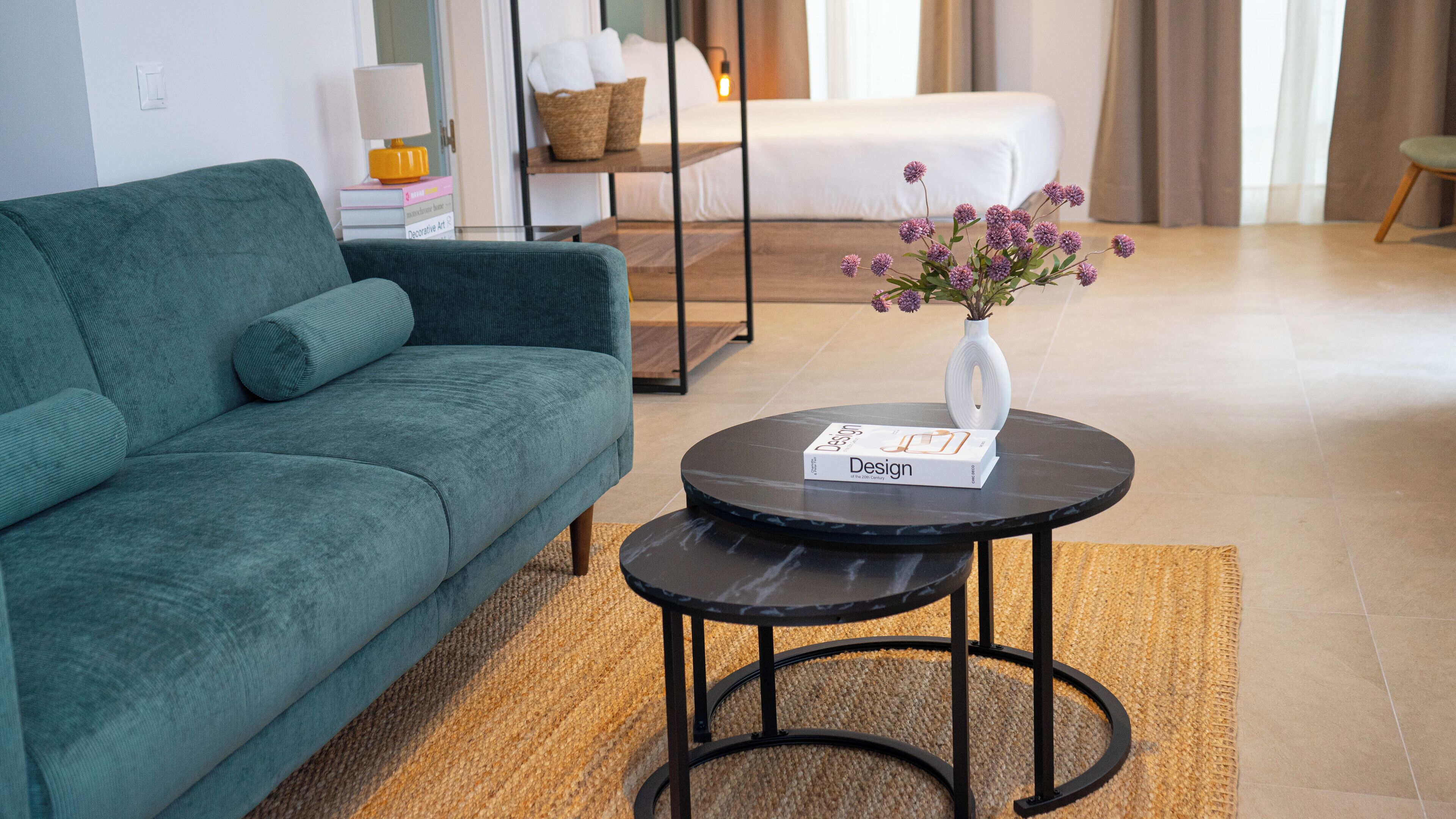 Uma sala de estar moderna com um sofá verde-água, uma mesa de centro redonda preta com um livro intitulado "Design" e um vaso com flores roxas.