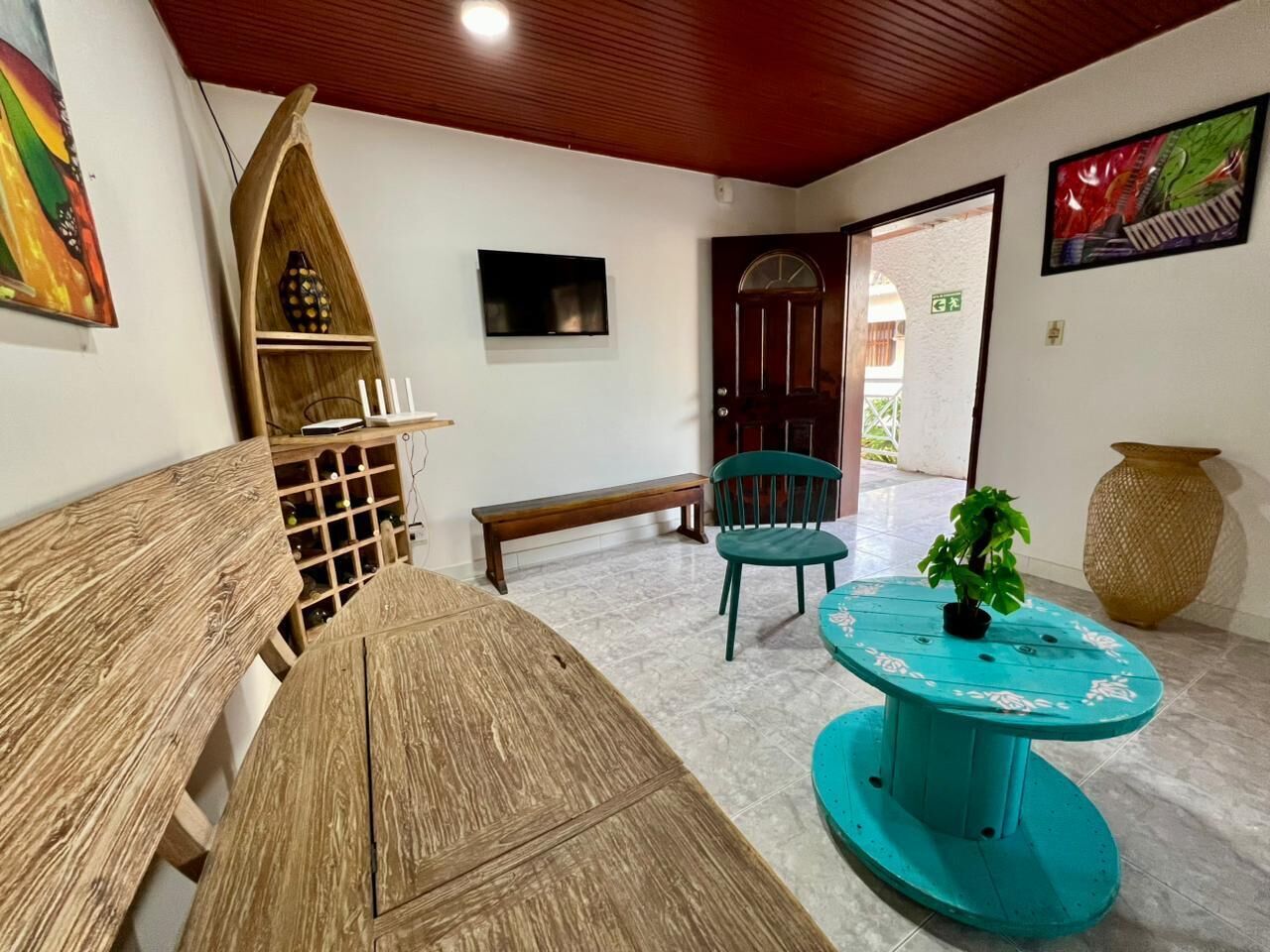 Quarto com um banco de madeira, uma mesa turquesa com uma planta, uma prateleira de madeira com garrafas e uma porta com sinalização de saída.