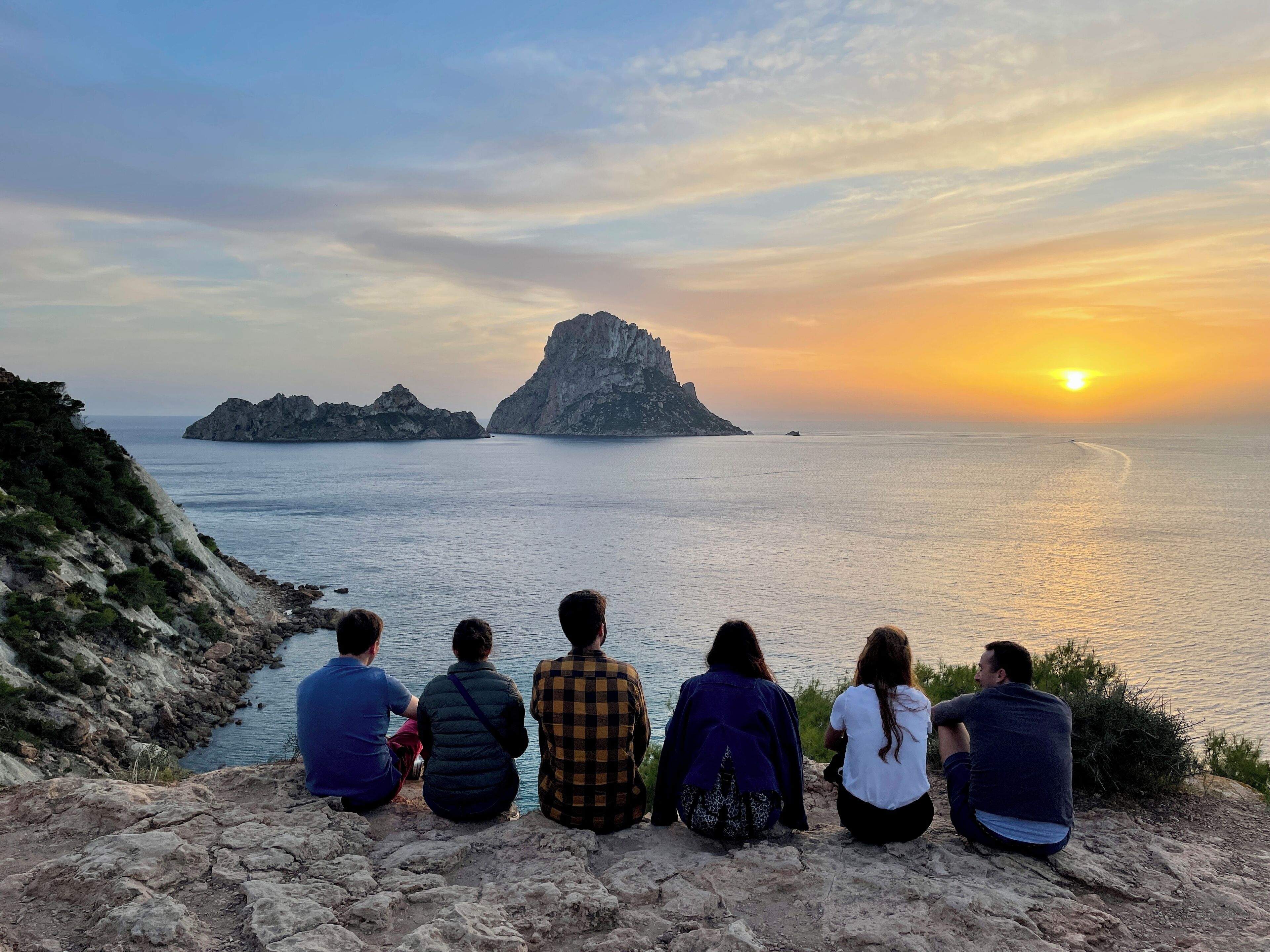 Um grupo de pessoas sentadas em um afloramento rochoso, com vista para um mar calmo e um pôr do sol ao fundo.