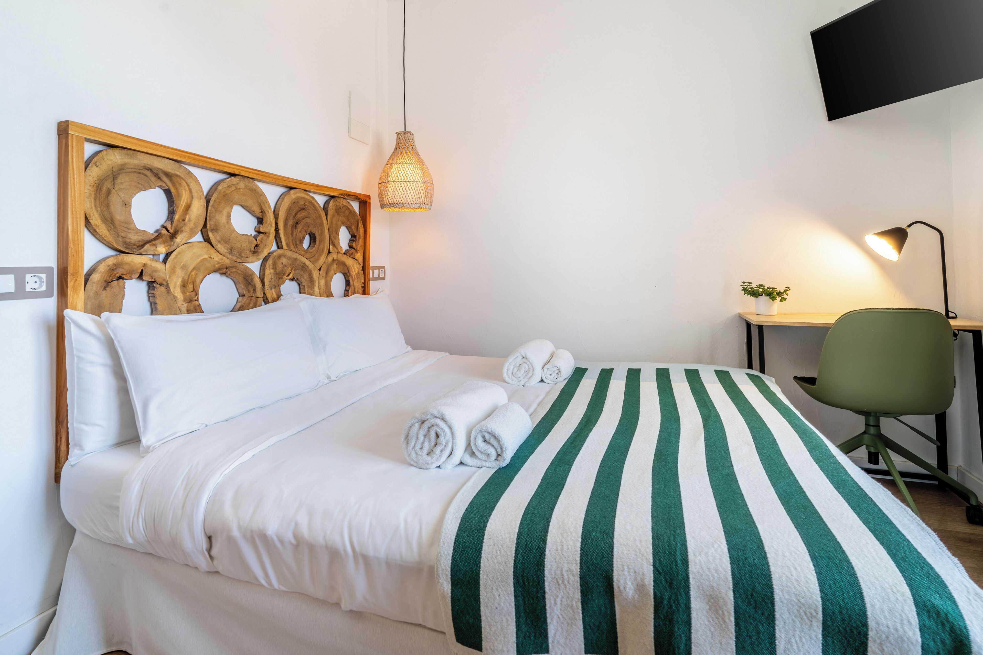 Cama bem arrumada com lençóis brancos e manta verde e branca listrada, cabeceira de madeira com recortes circulares, luminária pendente trançada, escrivaninha com cadeira e TV de tela plana na parede.