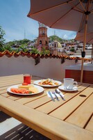 Mesa com pratos de comida, uma xícara e talheres, sob um guarda-sol, com vista para um horizonte urbano com edifícios e uma torre.