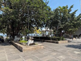 Uma praça pública ampla, com calçamento de pedra, árvores grandes e vista distante de um corpo d'água.