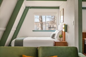Quarto com cama, mesa de cabeceira, abajur e janela com vista para árvores e edifícios.