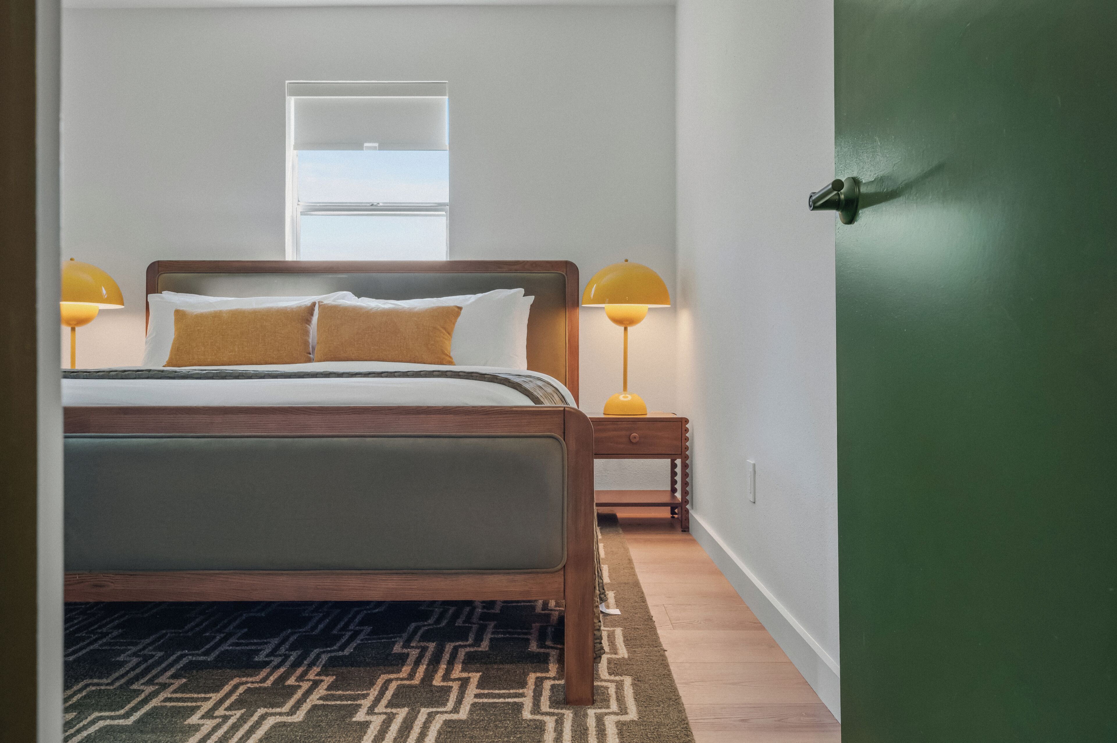 Quarto com cama, luminárias de cabeceira, janela e porta.