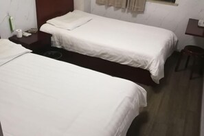 Quarto de hotel com duas camas de solteiro, uma mesinha, uma cadeira e uma mesa de cabeceira com abajur e alguns objetos.
