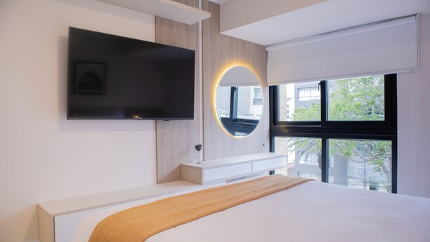 Quarto moderno com uma cama grande, televisão montada na parede, mesa de cabeceira e janela com vista para edifícios e árvores.