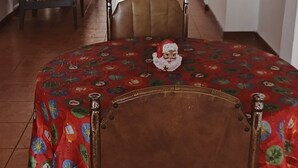 Uma sala de jantar com uma mesa redonda coberta por uma toalha vermelha com tema natalino, rodeada por quatro cadeiras.