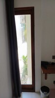 Um quarto com janela, cortina, prateleira de madeira e lixeira.