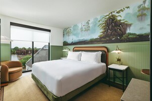 Quarto de hotel com uma cama grande, uma cadeira, uma mesa pequena e um mural na parede.