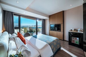 Quarto de hotel moderno com uma cama grande, TV, minibar e varanda com vista para a cidade.