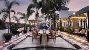 Uma área de refeições ao ar livre com uma mesa central rodeada por cadeiras, uma fonte decorativa e um pérgola com assentos.
