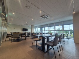 Uma sala de conferências espaçosa com uma mesa grande e cadeiras, uma TV de tela plana na parede e amplas janelas com vista para o exterior.