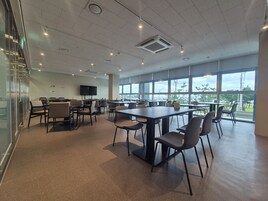 Sala de conferências com mesas, cadeiras, uma TV de tela plana e grandes janelas.