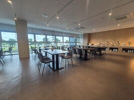 Amplo espaço interno com mesas e cadeiras dispostas para refeições, grandes janelas com vista para o exterior e um balcão com eletrodomésticos.