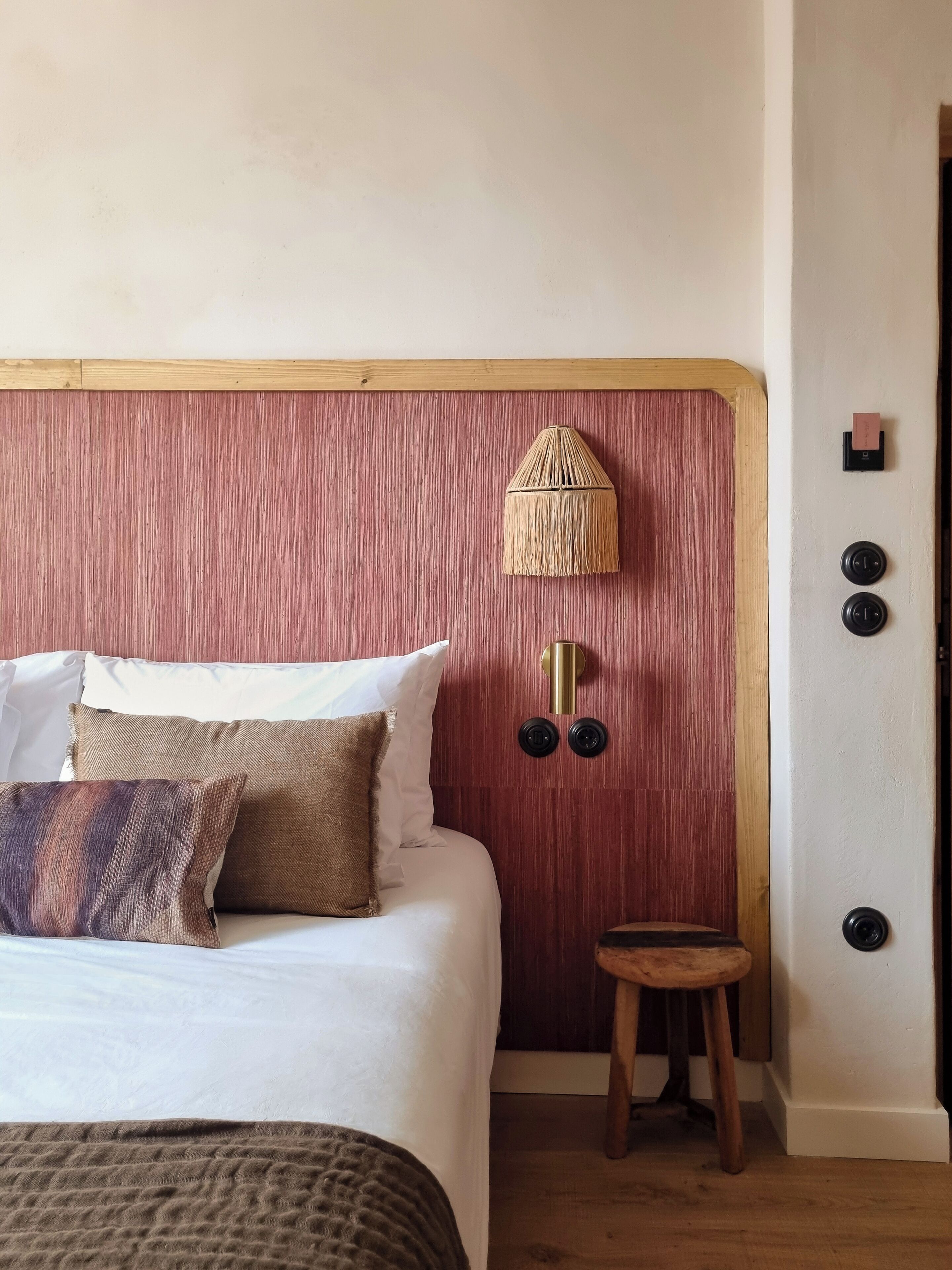 Cama bem arrumada com almofadas brancas e estampadas, cabeceira de madeira e um banquinho ao lado.