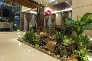Um lobby moderno de hotel com área de refeições, grandes plantas em vasos e um elemento decorativo de água.