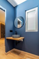 Casa de banho com parede azul, espelho redondo, pia com torneira preta e janela com moldura branca.