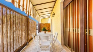 Uma área de estar coberta ao ar livre, com mesa, cadeiras, um vaso de planta e uma porta de madeira.