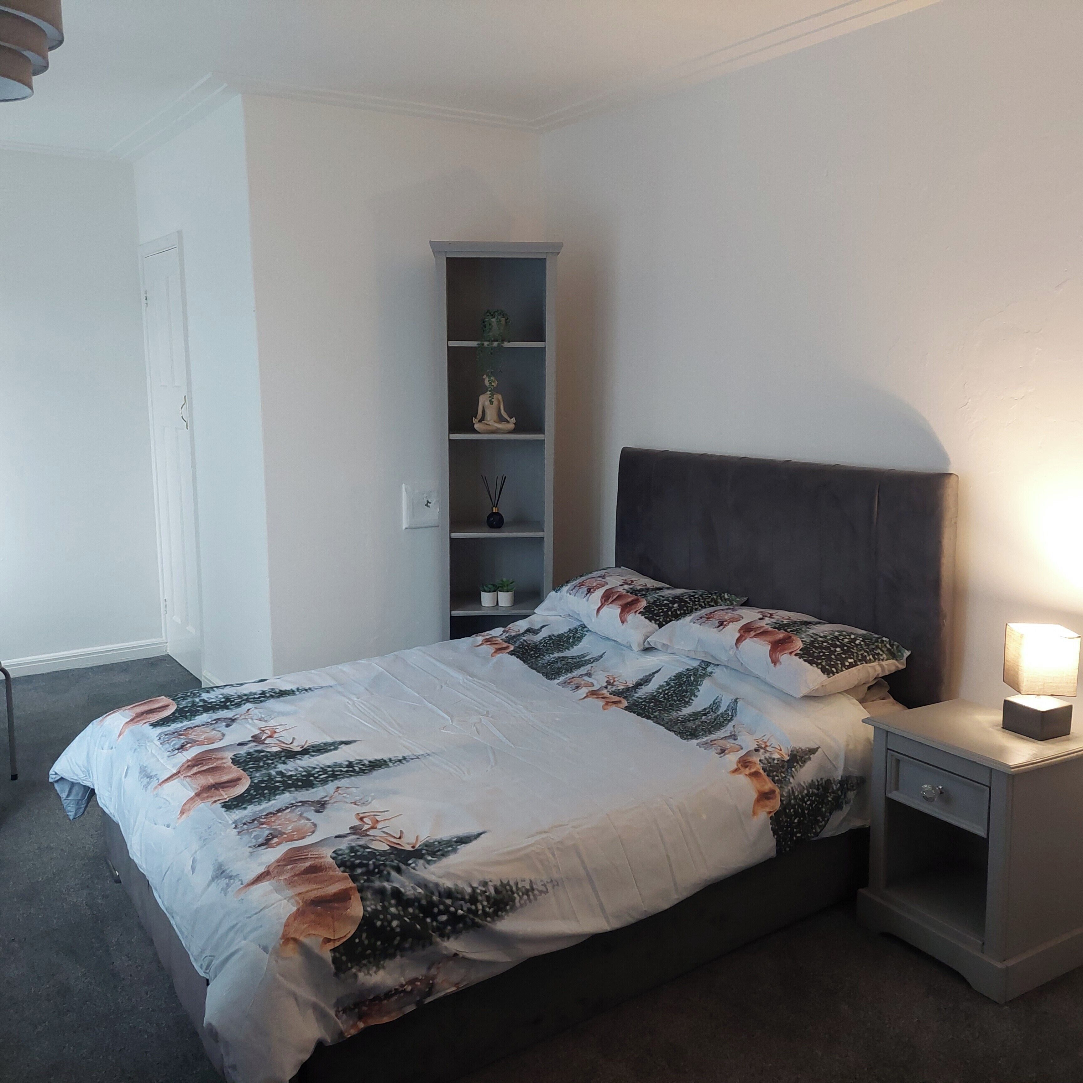 Quarto com uma cama, mesa de cabeceira, prateleira com objetos decorativos e uma porta.